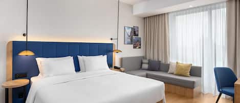 Quarto de hotel com uma cama grande, um sofá, uma cadeira, uma escrivaninha e uma janela com cortinas.