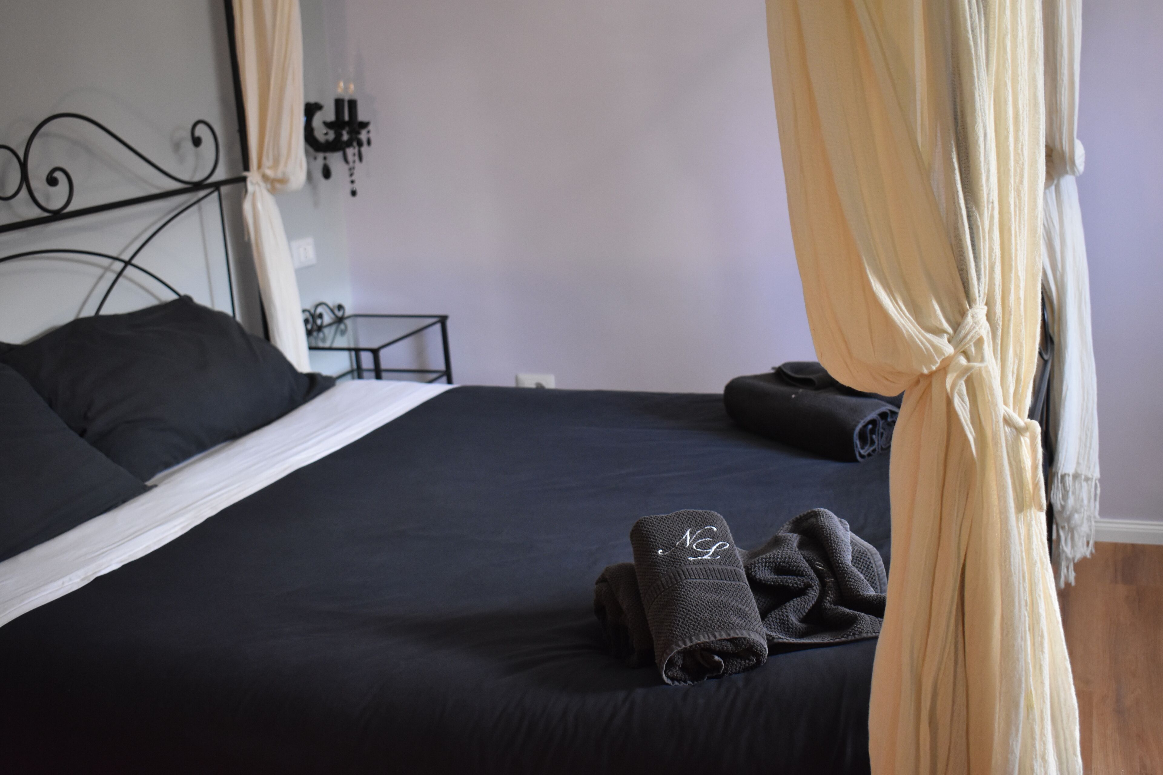 Cama arrumada com um edredom preto, duas toalhas enroladas e um dossel com cortina branca.