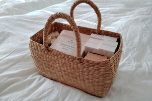 Cesto de vime com alças contendo vários objetos sobre um cobreleiro branco.