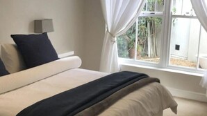 Cama bem arrumada com roupa de cama branca e escura, abajur e janela com cortinas.