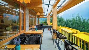 Restaurante com mesas e cadeiras de madeira, janelas amplas e um corrimão amarelo com vista para o verde.