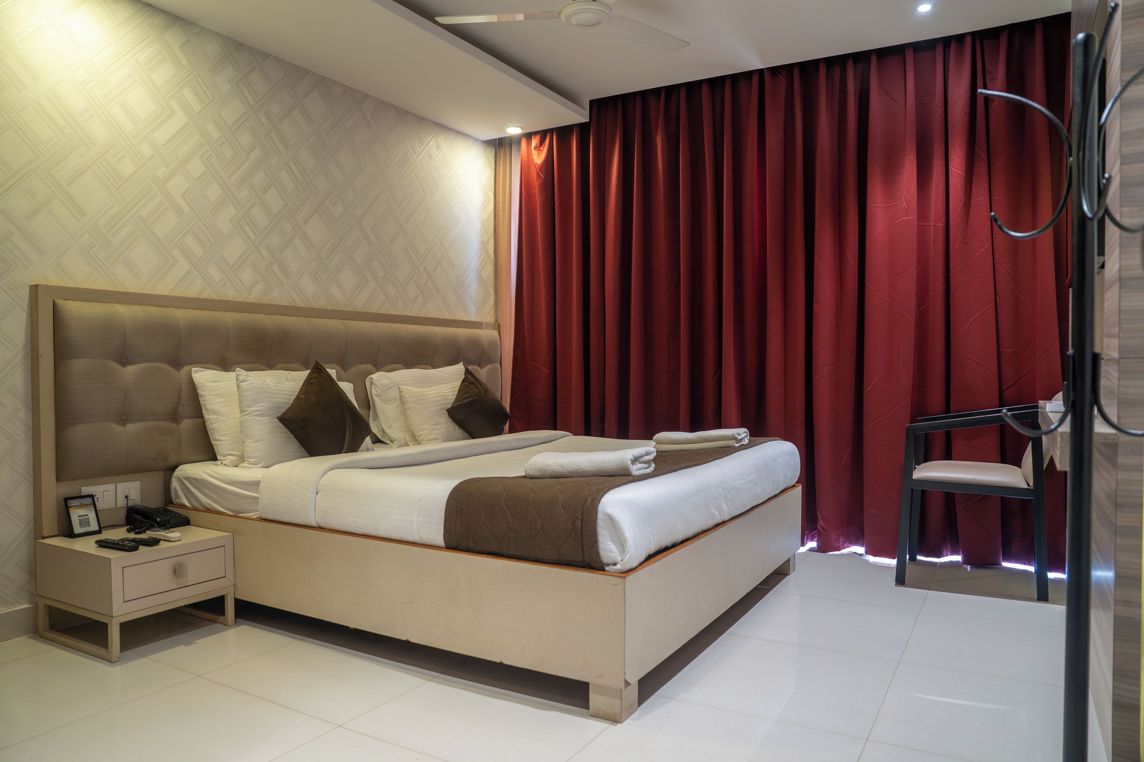 Quarto de hotel moderno com uma cama grande, mesa de cabeceira com telefone e controles remotos, uma cadeira e cortina vermelha.