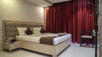 Quarto de hotel moderno com uma cama grande, mesa de cabeceira com telefone e controles remotos, uma cadeira e cortina vermelha.