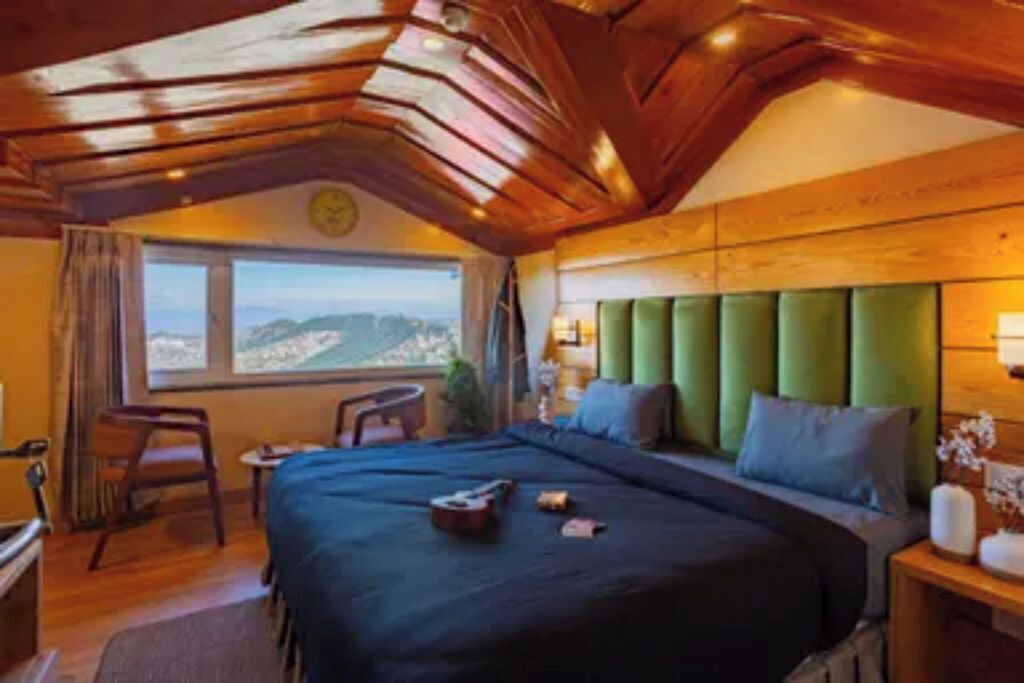 Quarto com cama grande, teto de madeira, vista pela janela e uma pequena mesa com uma cadeira.