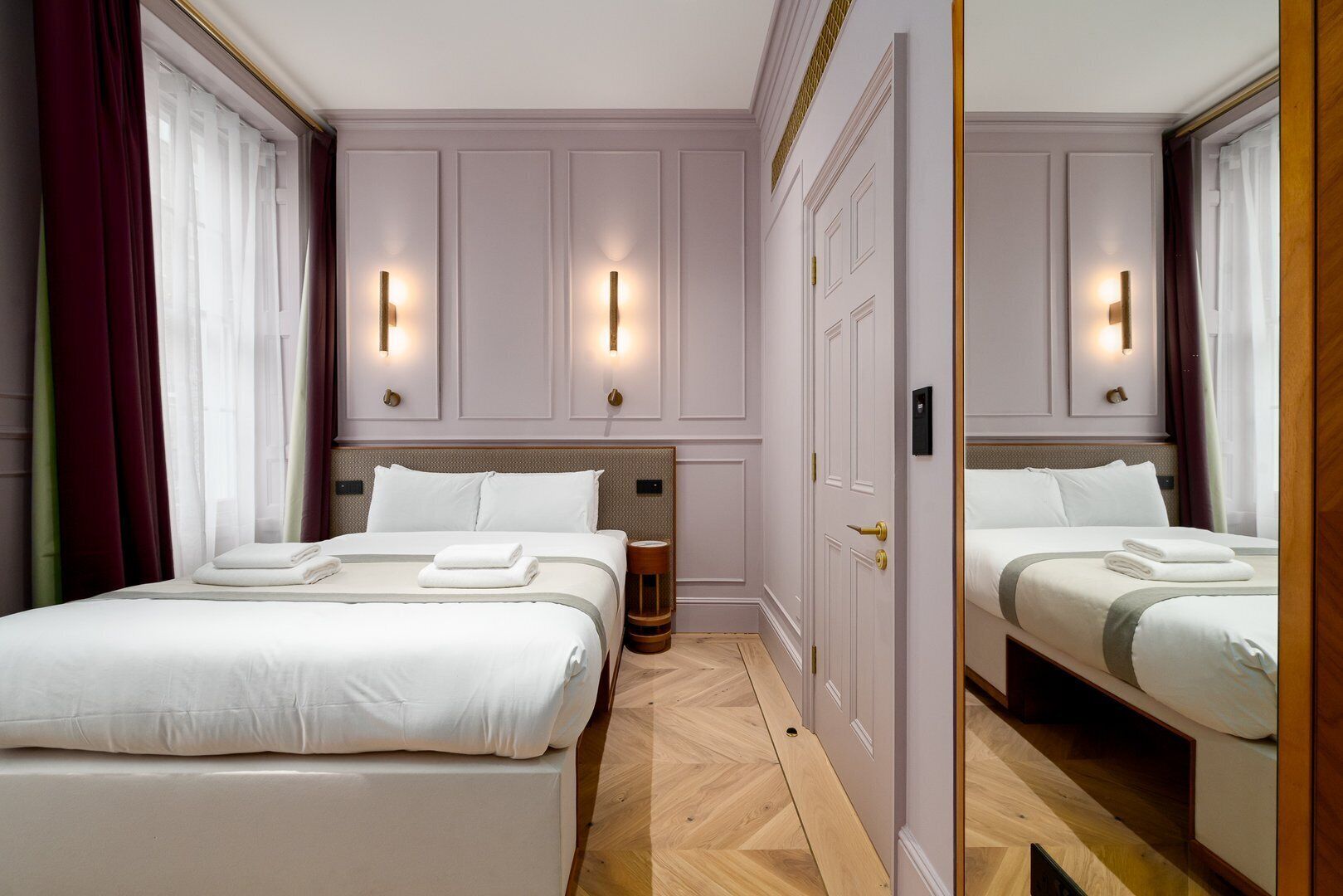 Quarto de hotel com duas camas, piso de madeira e um espelho refletindo o ambiente.