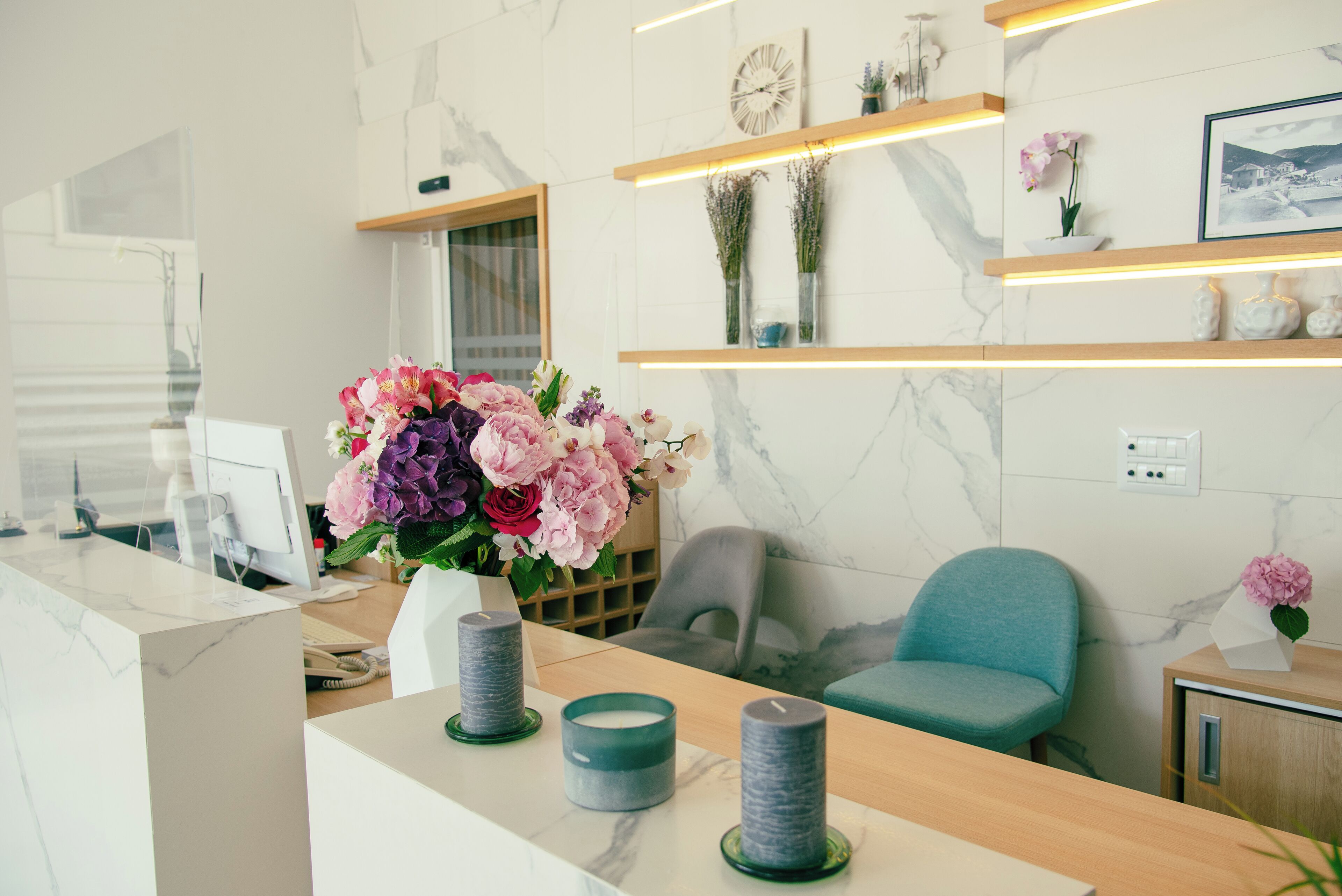 Uma área de recepção com um balcão de mármore, uma cadeira azul e uma estante com objetos decorativos e um vaso de flores.