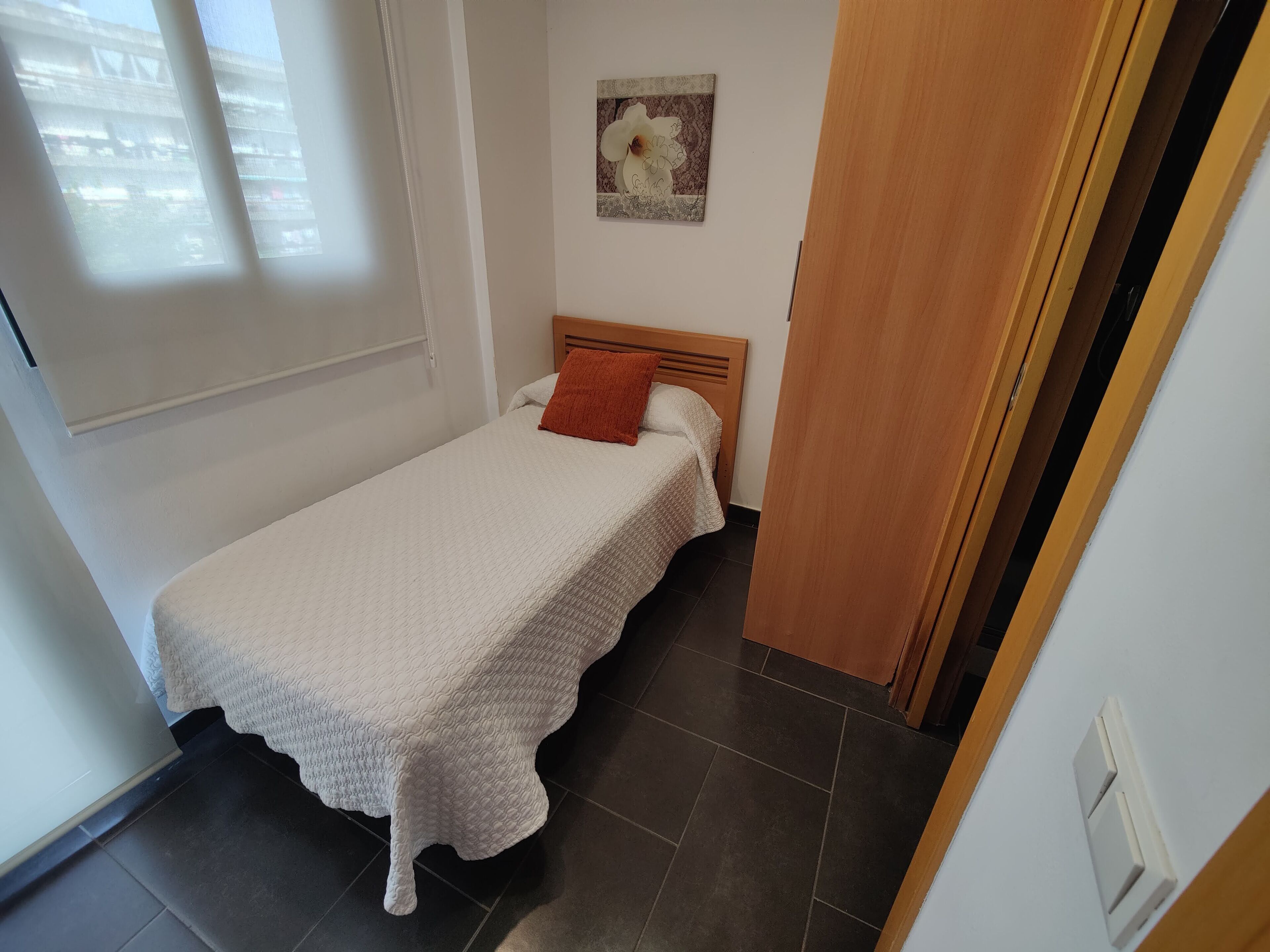 Cama pequena e bem arrumada com colcha branca e um único travesseiro laranja em um quarto com chão de azulejos escuros e paredes brancas.