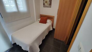 Cama pequena e bem arrumada com colcha branca e um único travesseiro laranja em um quarto com chão de azulejos escuros e paredes brancas.