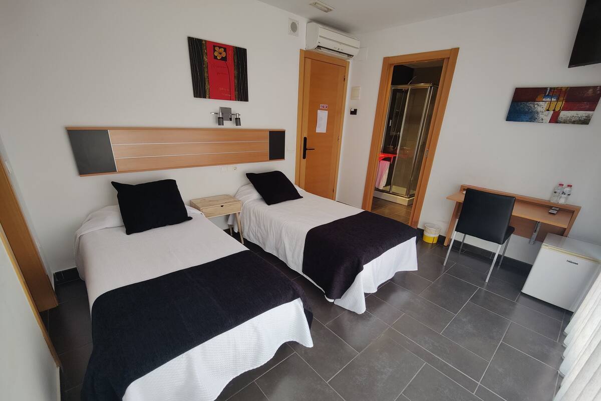 Quarto de hotel com duas camas, uma escrivaninha, uma cadeira e um frigorífico.