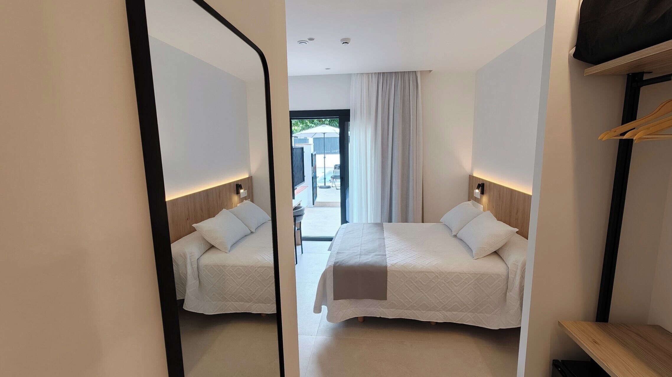 Quarto de hotel com duas camas, um espelho, cabeceira de madeira, uma mesinha e vista para o exterior.