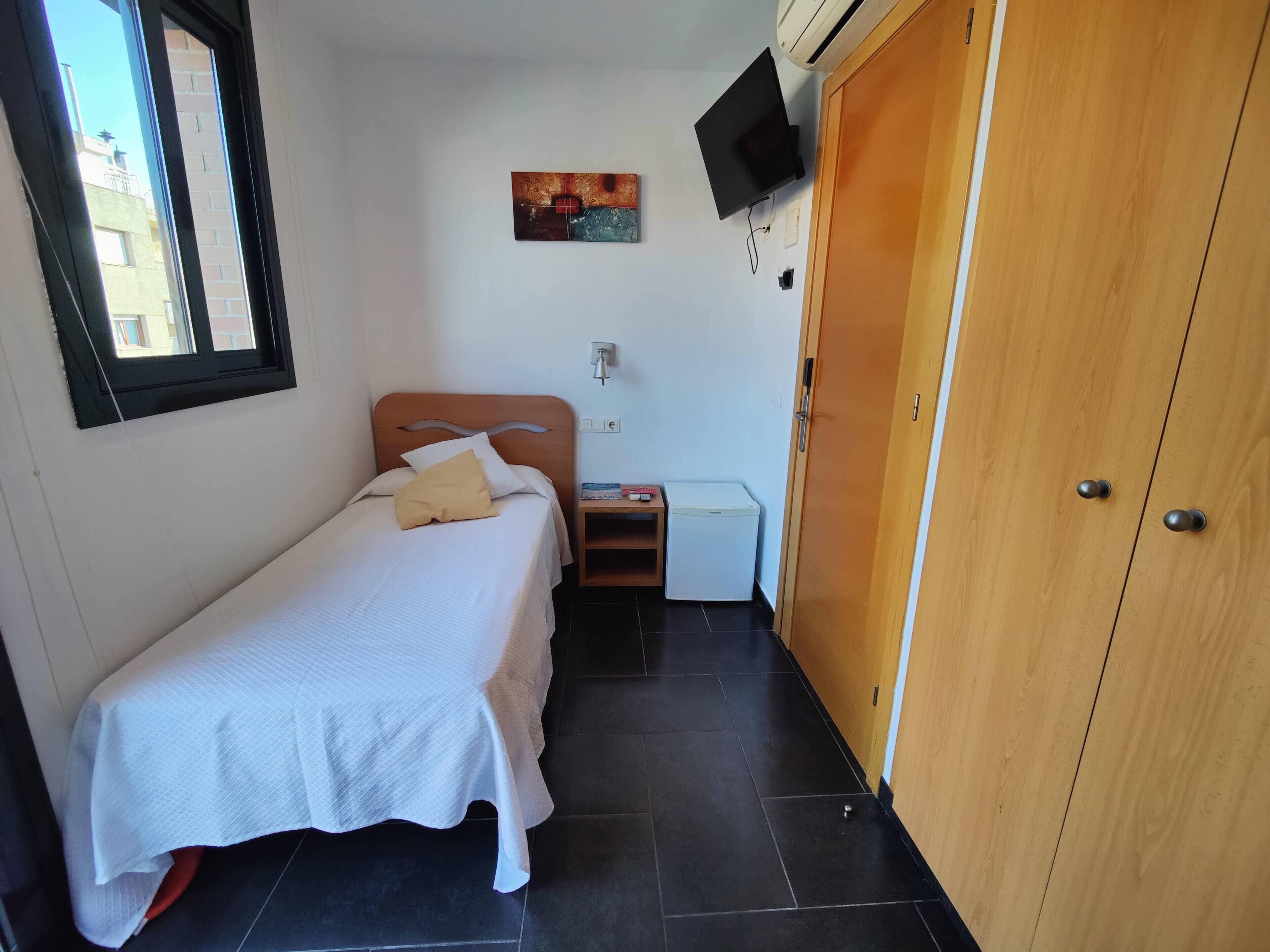 Um quarto pequeno e arrumado, com uma cama, mesa de cabeceira, TV fixada na parede e um guarda-roupa.