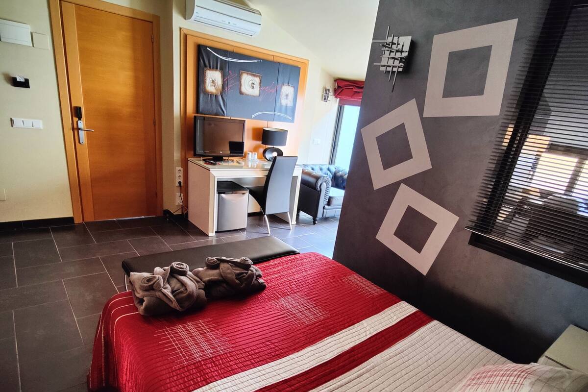 Quarto de hotel com uma cama, uma escrivaninha e uma cadeira. Há uma porta, uma janela com persianas e uma parede com padrão geométrico.