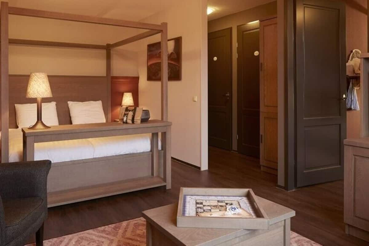 Uma cama de dossel com luminárias, uma mesinha com bandeja, uma cadeira e uma televisão sobre um aparador.
