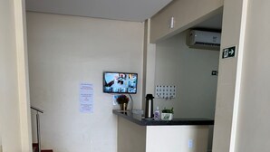 Uma área de recepção com um banco de madeira, um balcão, uma televisão fixada na parede e um mural de avisos.