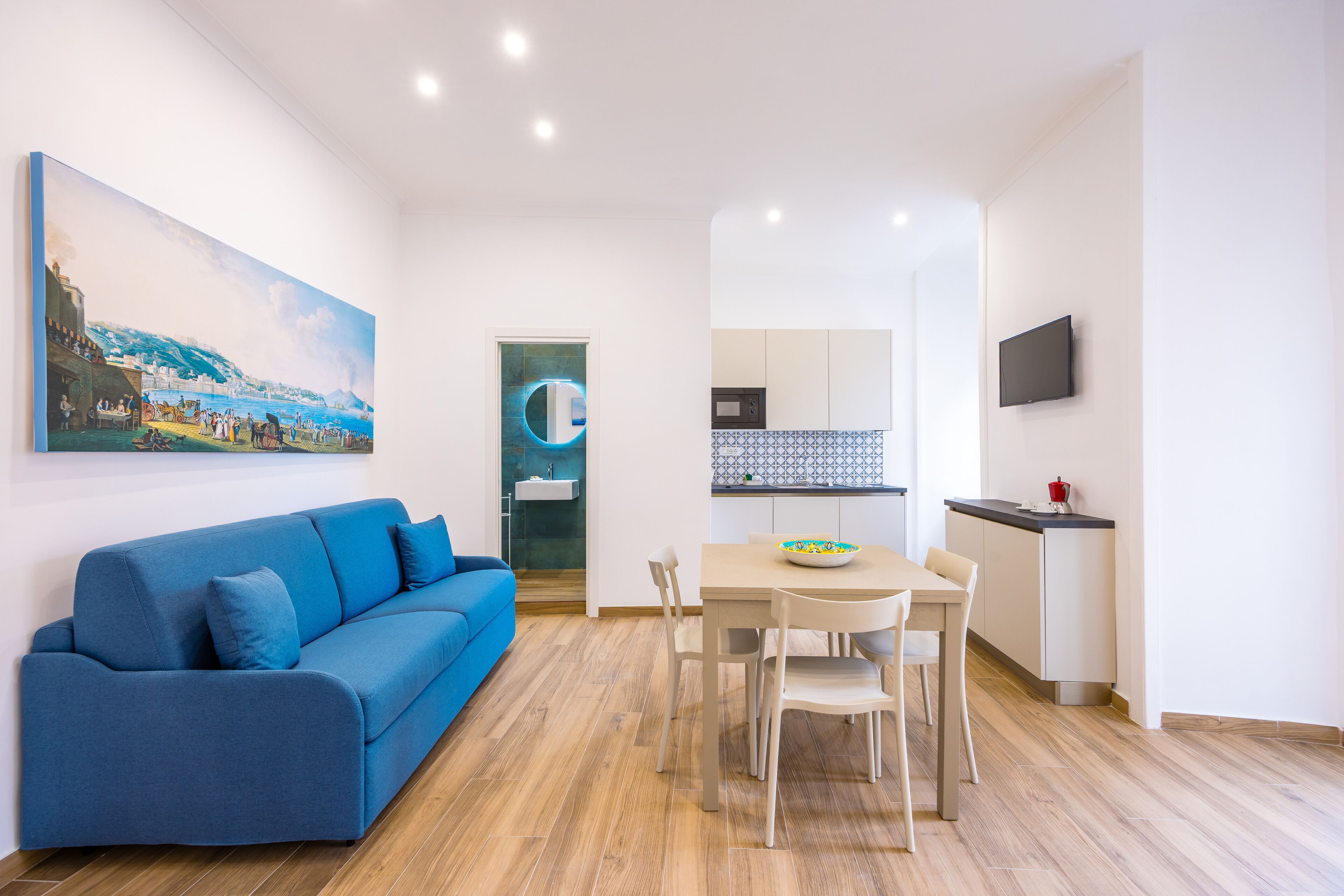 Uma sala de estar moderna com um sofá azul, uma mesa de jantar com cadeiras, uma área de cozinha com televisão e um quadro na parede.