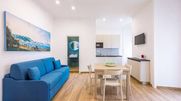 Uma sala de estar moderna com um sofá azul, uma mesa de jantar com cadeiras, uma área de cozinha com televisão e um quadro na parede.