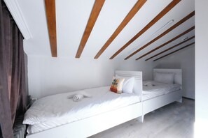 Uma cama de solteiro com cabeceira branca e um travesseiro com padrão de folha. O quarto possui vigas de madeira no teto e uma parede branca.