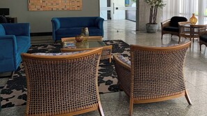 Um saguão de hotel com design moderno, apresentando uma grande obra de arte abstrata na parede, uma área de estar com cadeiras de vime e um sofá azul, e uma grande janela com cortinas leves.