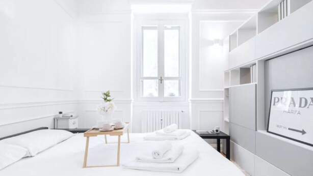 Quarto de hotel moderno com uma cama grande, uma mesa de cabeceira com um vaso e uma janela com persianas.