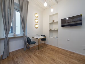 Quarto moderno e bem iluminado com uma mesa de jantar, duas cadeiras, uma TV fixada na parede e uma janela com cortinas.