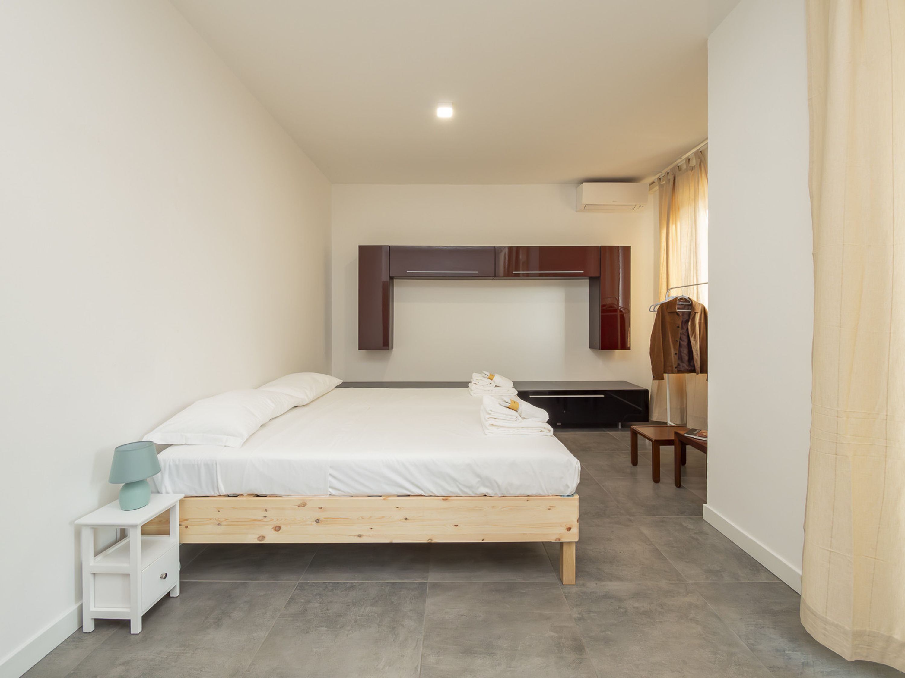 Quarto com cama, mesa de cabeceira, cómoda de madeira, cabideiro e luminária no teto.