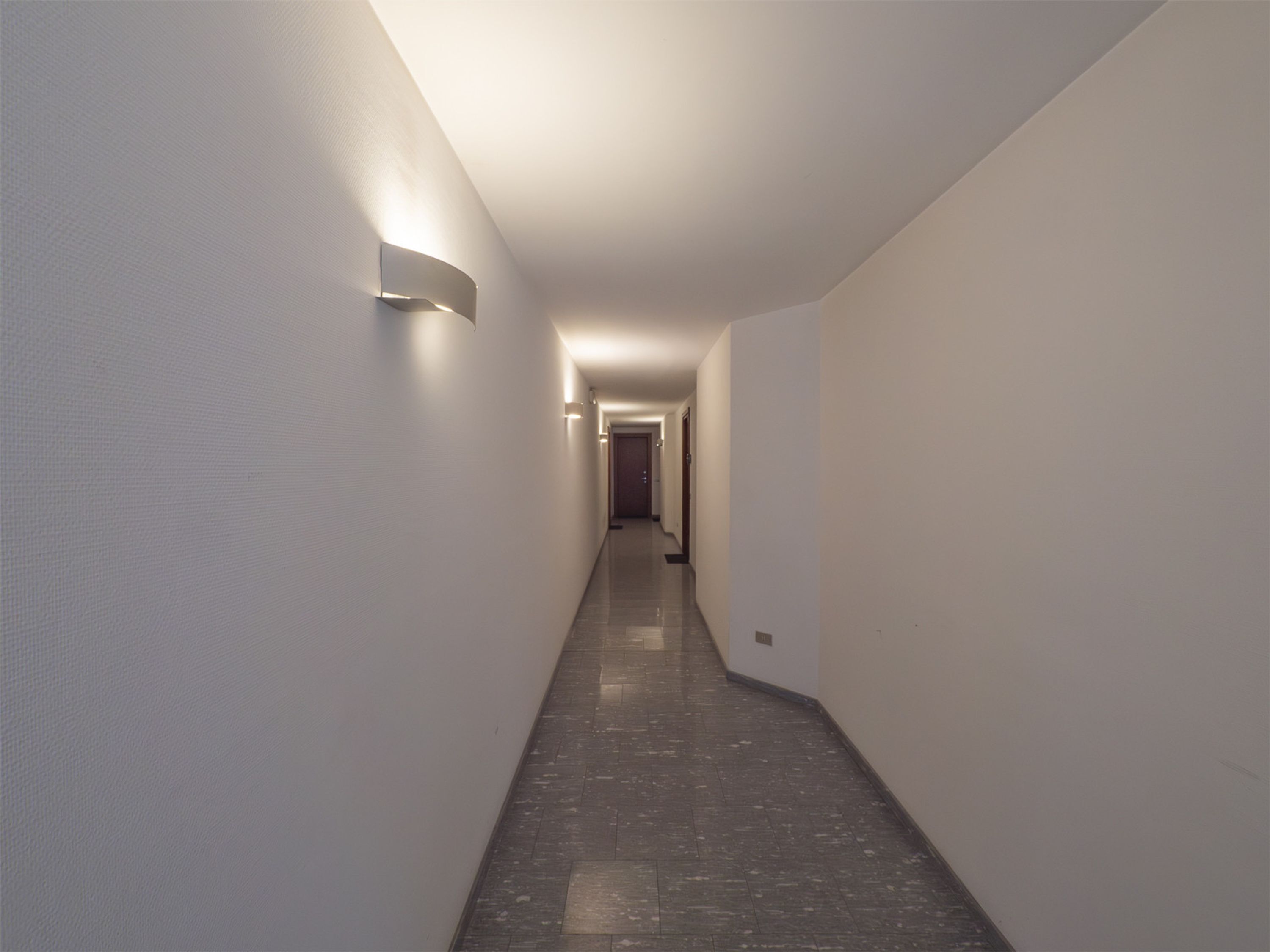 Um corredor estreito, bem iluminado, com piso de azulejos e paredes brancas.