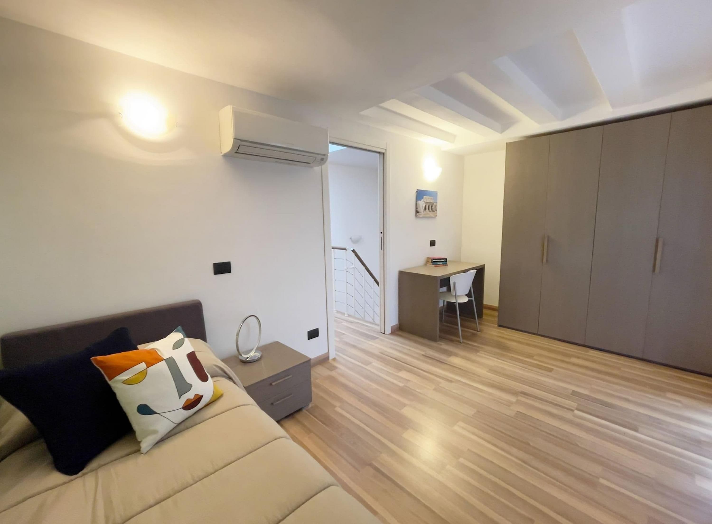 Quarto moderno com cama, mesa de cabeceira e escrivaninha com cadeira. Há um guarda-roupa e uma porta que dá para outro cômodo.