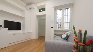 Uma sala de estar moderna com um sofá cinzento, uma televisão de ecrã plano fixada num móvel branco, uma janela com vista para edifícios e uma planta de cacto num vaso decorativo.