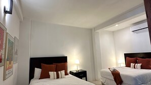 Quarto de hotel com duas camas, uma mesa de cabeceira com abajur e um aparelho de ar condicionado na parede.