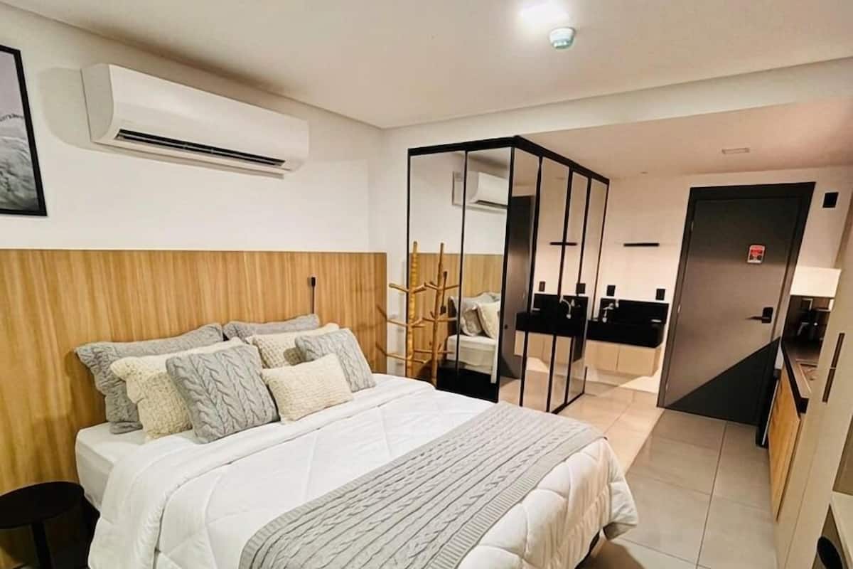 Quarto moderno com uma cama grande, cabeceira de madeira, divisória de vidro, banheiro com pia e espelho, e uma porta que dá para outro espaço.