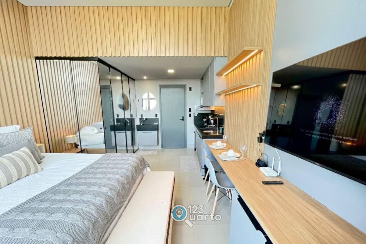 Quarto de hotel moderno com cama, escrivaninha com TV, área de cozinha e banheiro.