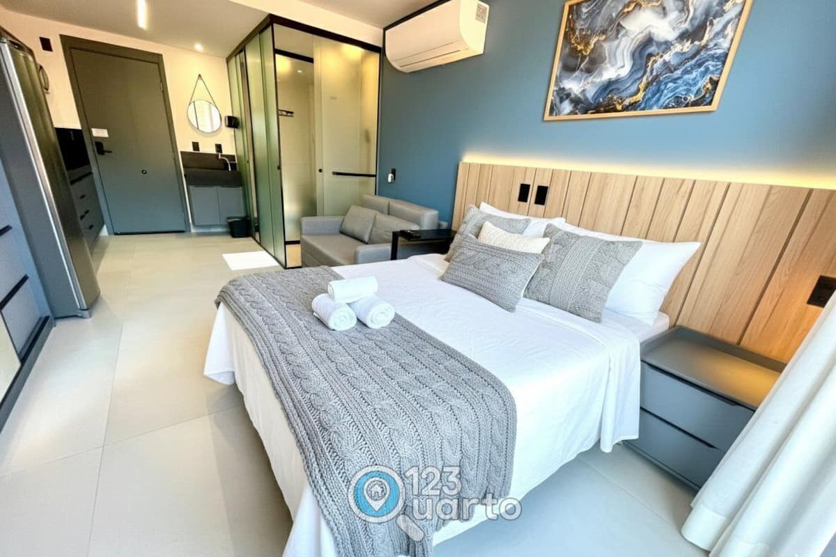 Quarto de hotel moderno com uma cama grande, cabeceira de madeira, box de chuveiro de vidro e um quadro na parede.