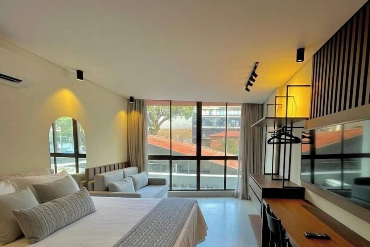 Quarto de hotel moderno com uma cama grande, área de estar, televisão e vista da cidade.
