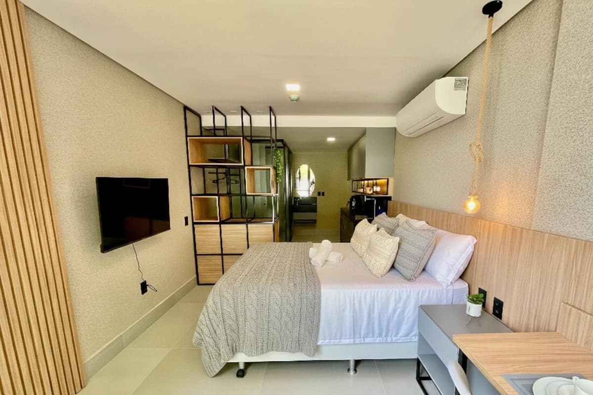 Quarto moderno com cama, mesa de cabeceira, televisão e vista para outro cômodo.