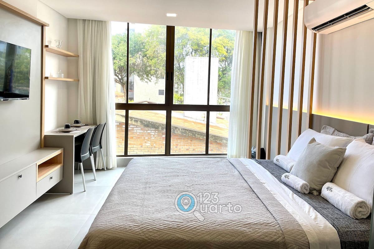 Quarto de hotel com cama, escrivaninha, cadeira, televisão e uma grande janela.
