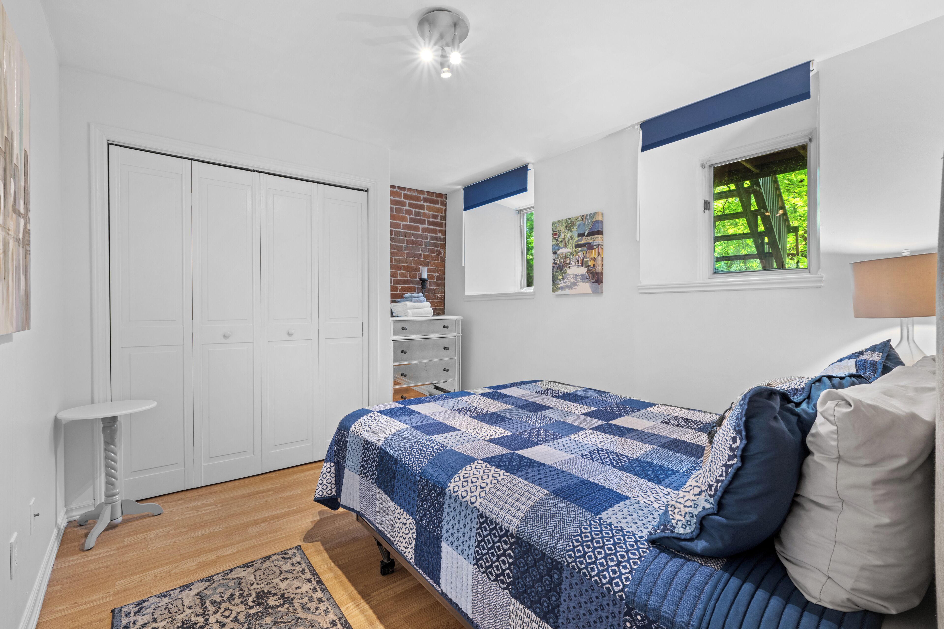 Quarto com cama, penteadeira, guarda-roupa, janela com vista para o verde e luminária no teto.