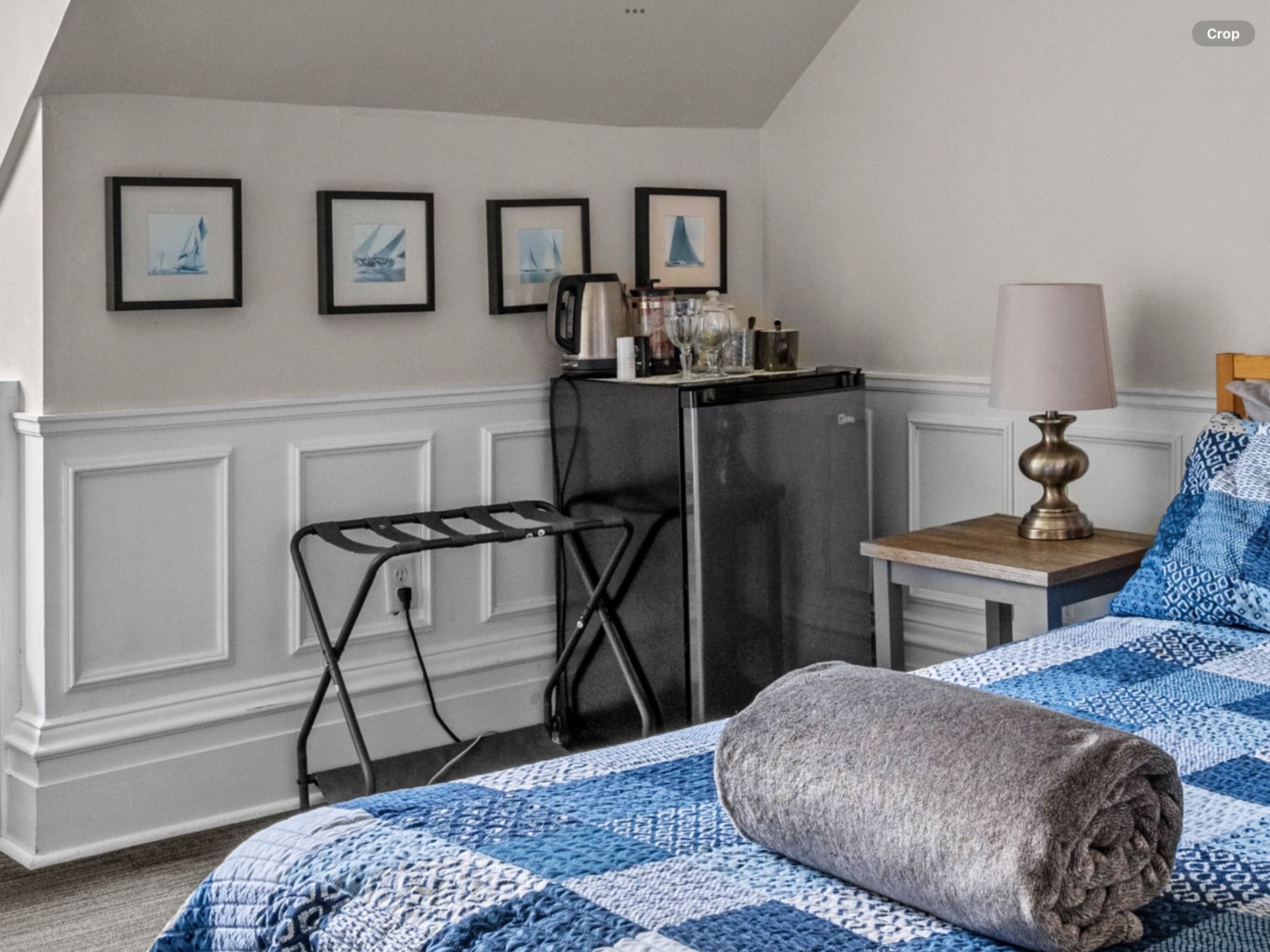 Quarto com cama, uma mesinha de cabeceira com luminária, geladeira e tábua de passar roupa fixada na parede.