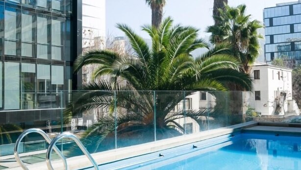 Uma piscina no terraço com águas azul-claras, cercada por palmeiras e edifícios modernos.