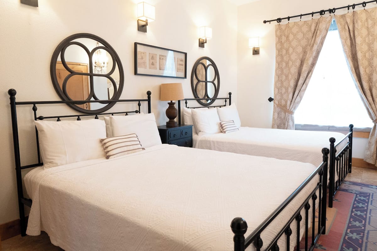 Quarto de hotel com duas camas, uma mesa de cabeceira, luminárias fixadas na parede e uma janela com cortinas.