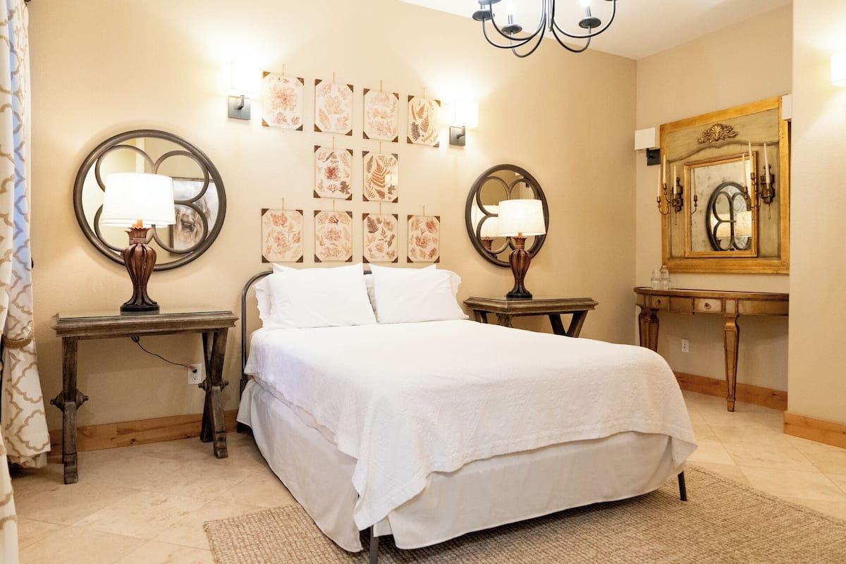 Quarto de hotel com uma cama, duas mesinhas de cabeceira com abajures, um espelho grande e uma obra de arte floral na parede.