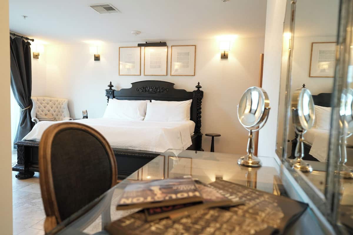 Quarto de hotel bem arrumado, com uma cama grande, uma cadeira, uma mesa com revistas e um espelho.