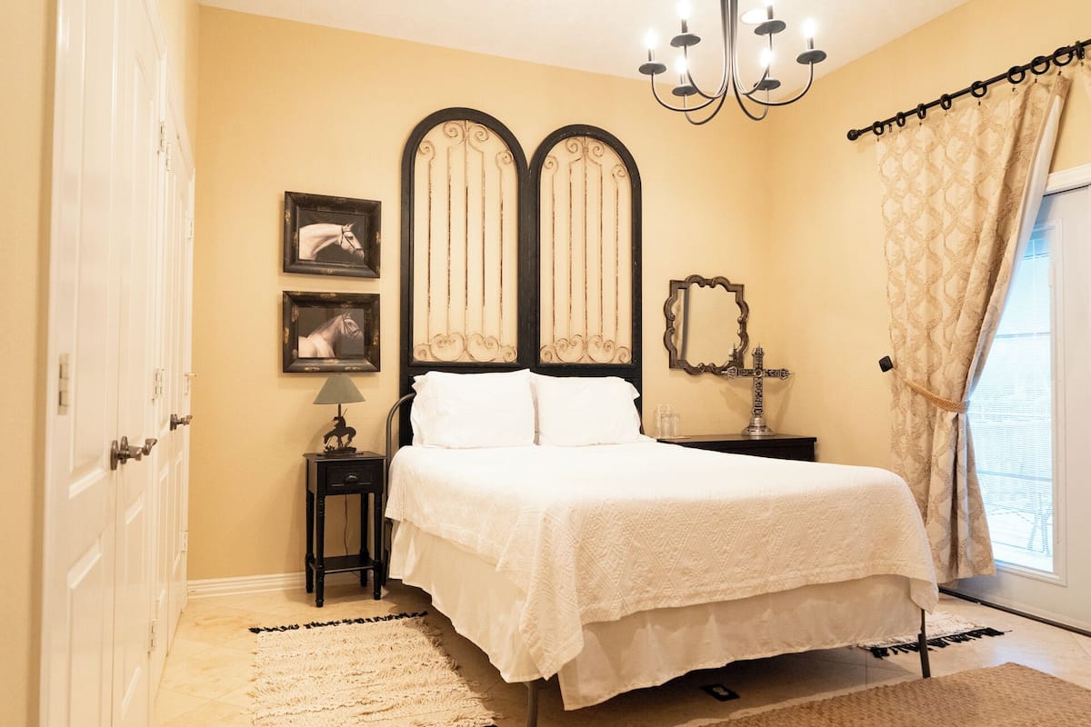 Um quarto com cama, mesa de cabeceira, luminária, lustre e janela com cortinas.