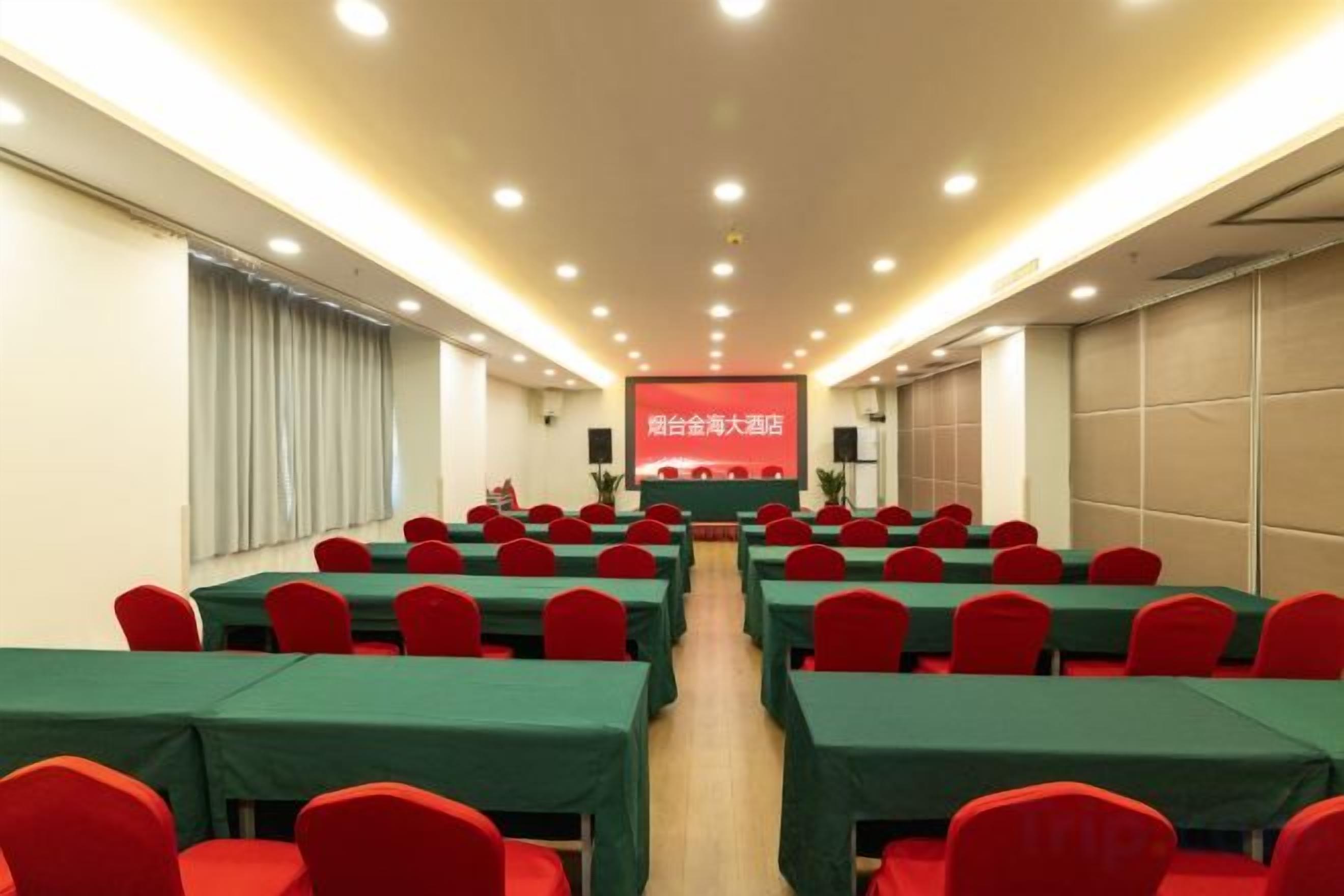 Sala de conferências com mesas e cadeiras dispostas em fileiras, uma tela de projeção e um palco na frente.