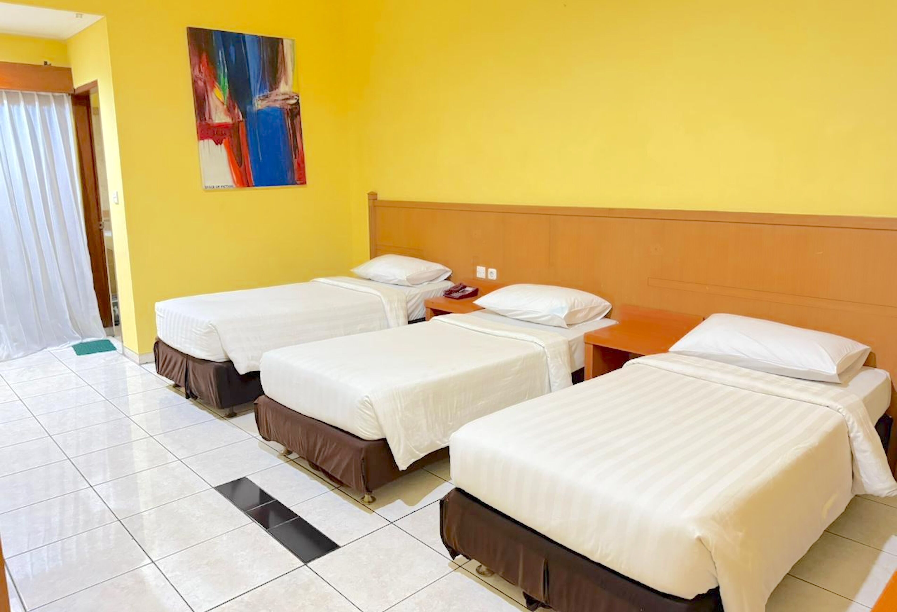 Quarto de hotel com três camas, uma cabeceira de madeira e uma pintura abstrata colorida na parede.