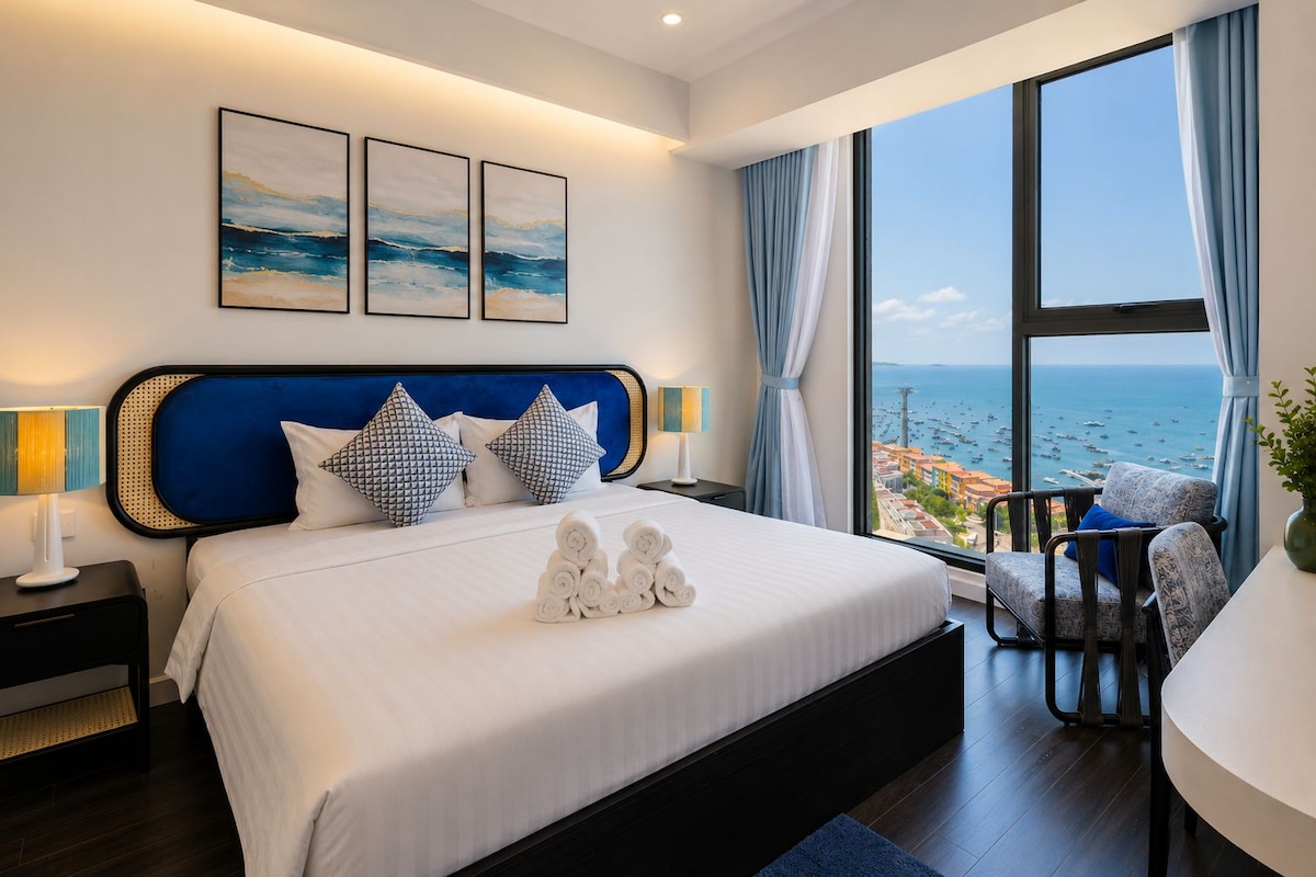 Quarto de hotel moderno com uma cama grande, duas luminárias laterais, vista para o mar e um espaço de estar com uma mesa.
