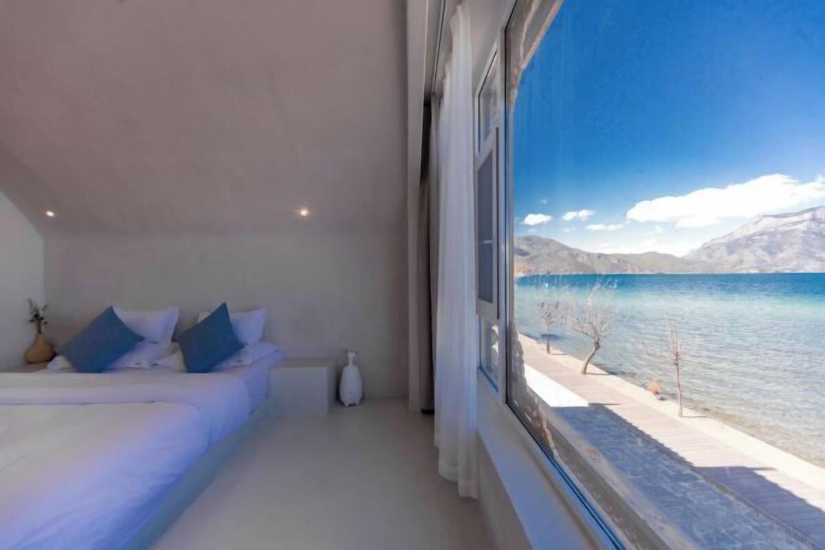 Quarto com cama, almofadas, mesa de cabeceira, janela e vista para um lago.