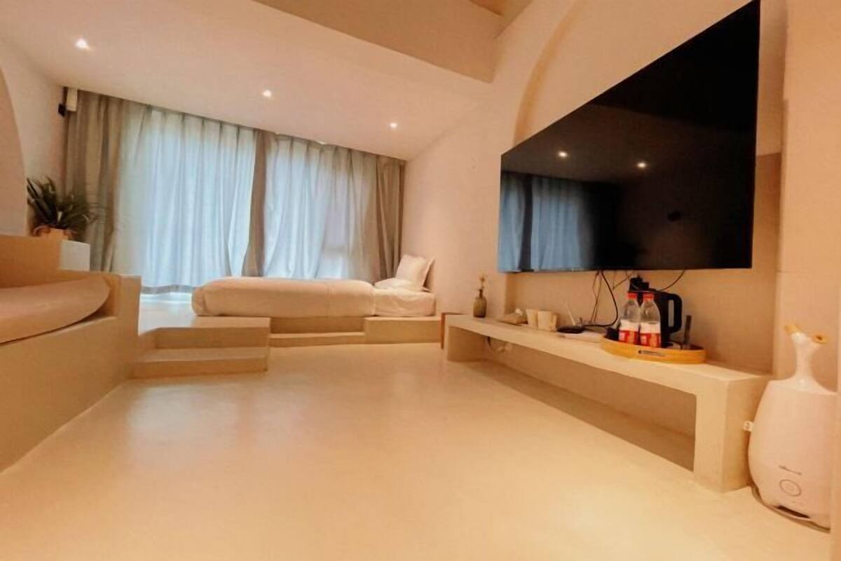 Quarto de hotel moderno com uma cama grande, televisão de tela plana na parede, um pequeno espaço de estar e mesa de cabeceira com garrafa e copos.