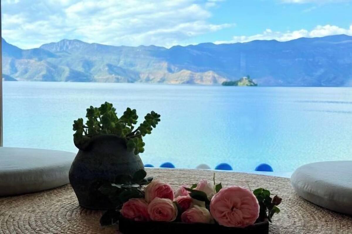 Vista de um lago com montanhas ao fundo, uma planta em vaso e uma tigela com flores cor-de-rosa sobre uma superfície texturizada.
