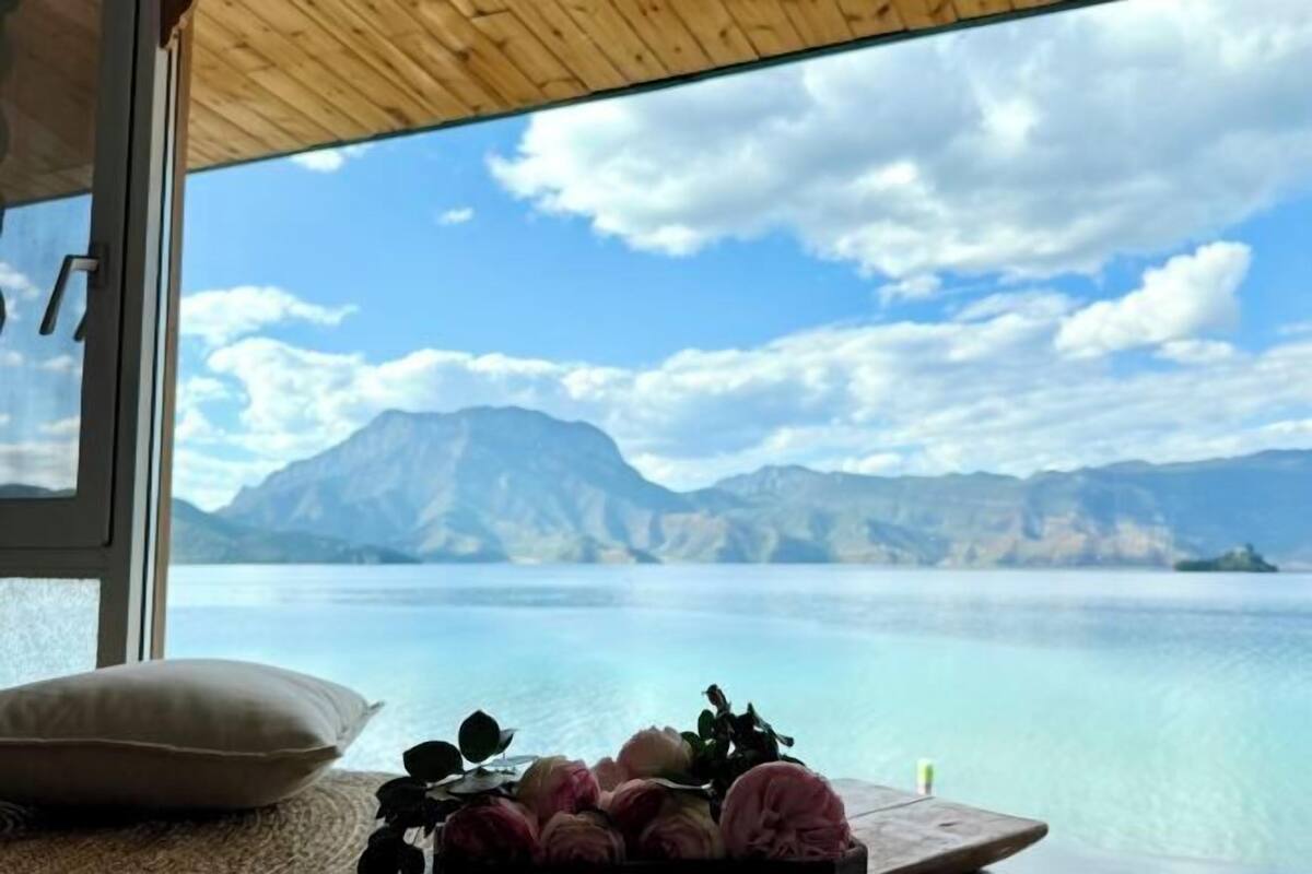 Quarto com vista para um lago e montanhas, teto de madeira, janela e mesa com flores.