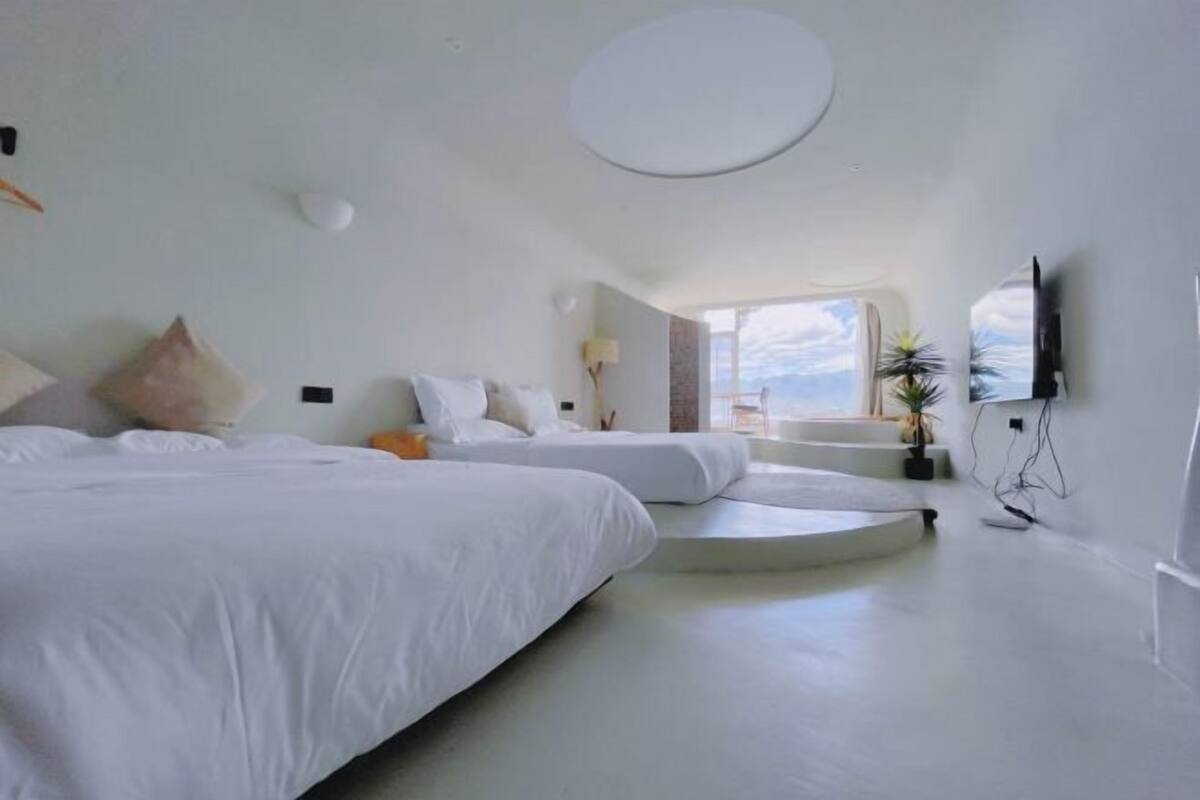 Quarto de hotel moderno com duas camas grandes, uma luminária redonda no teto, uma TV fixada na parede e vista do céu pela janela.